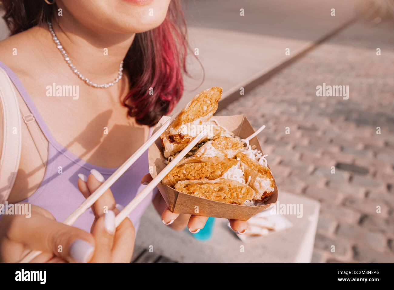 Happy girl eating spring rolls from takeaway paper box using chopsticks ...
