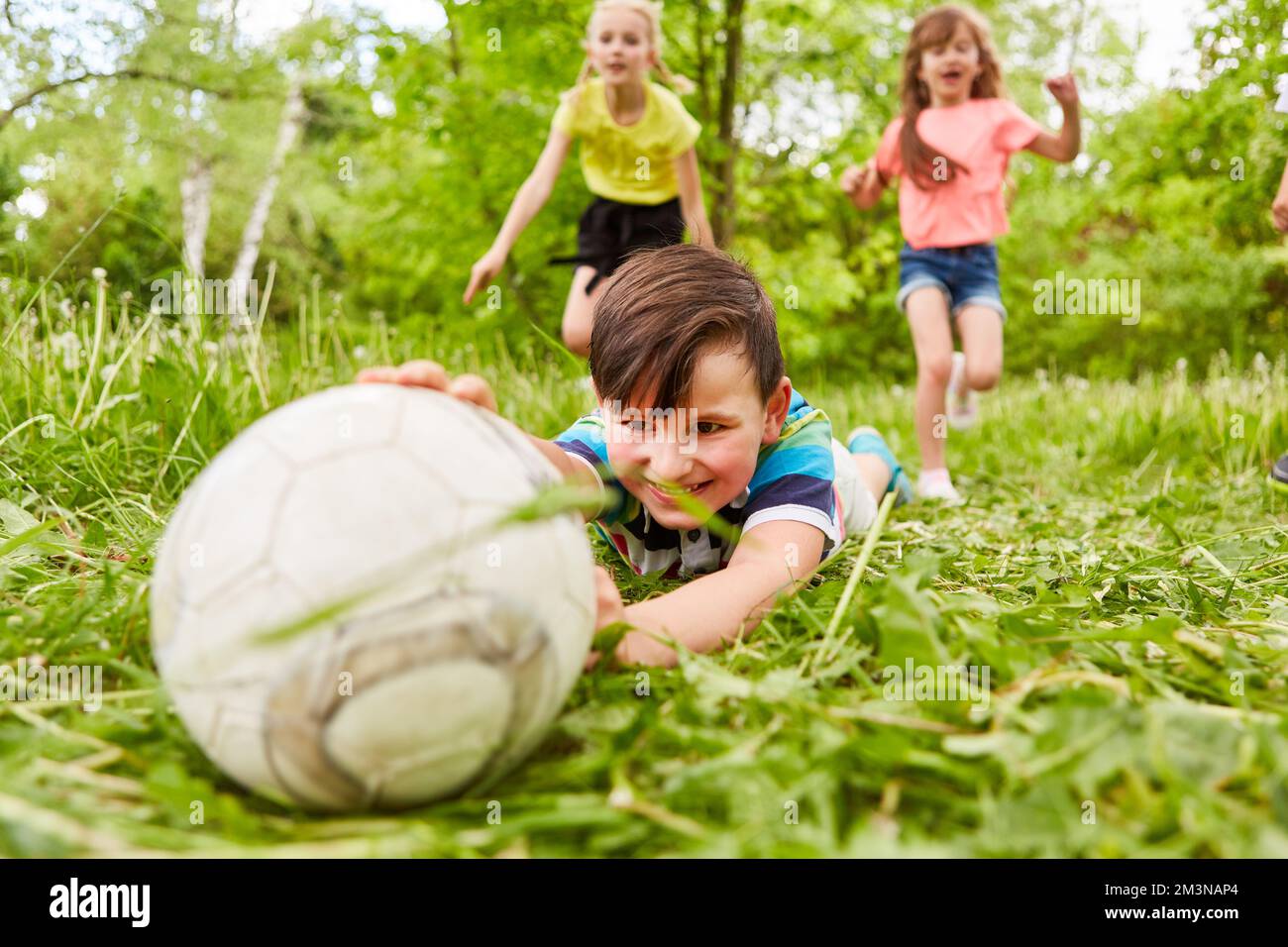 Happy boy playing football with friends running in background at garden during holiday Stock ...