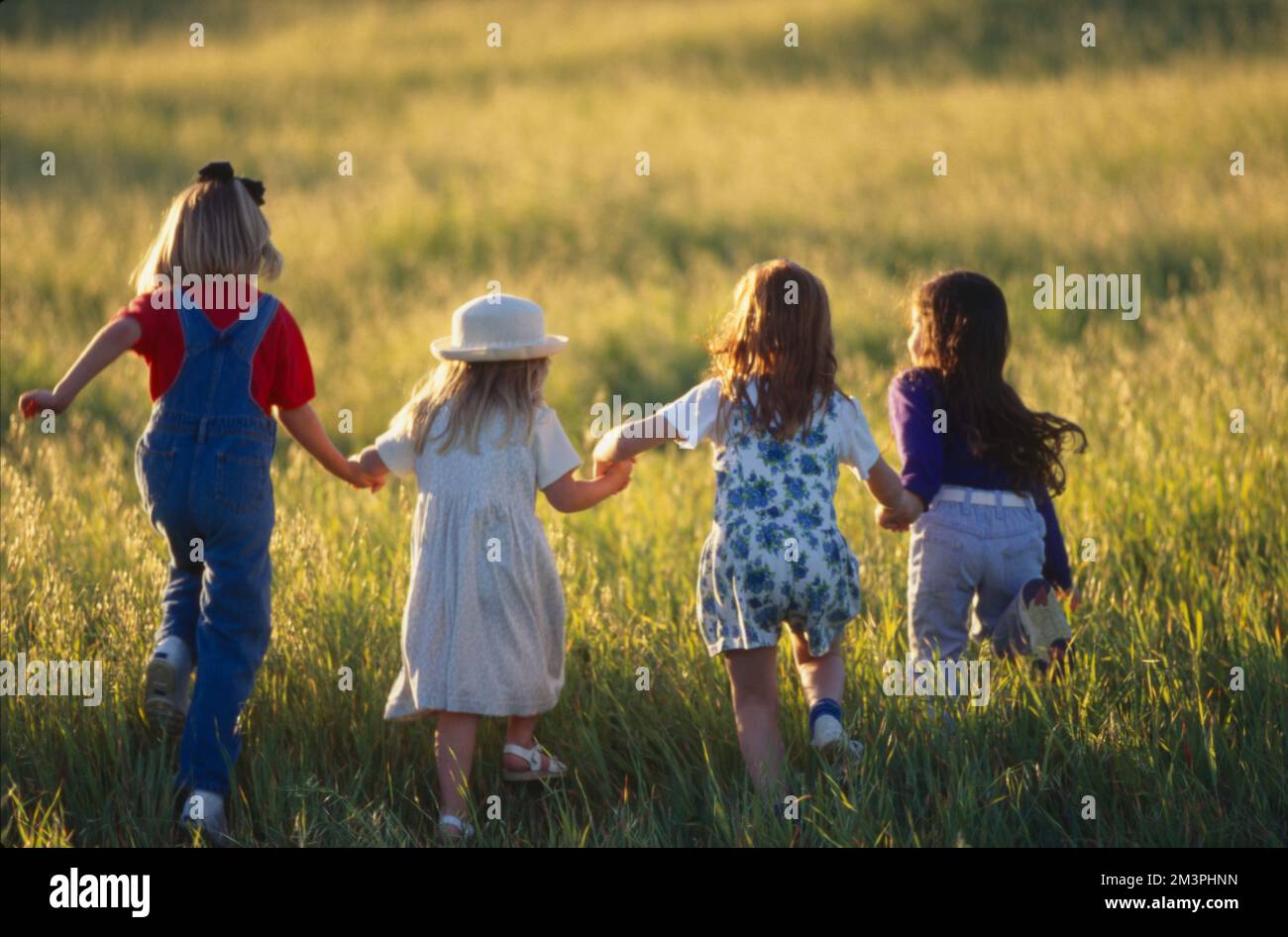 Four children pictured from behind romping thru the grass holding hands ...