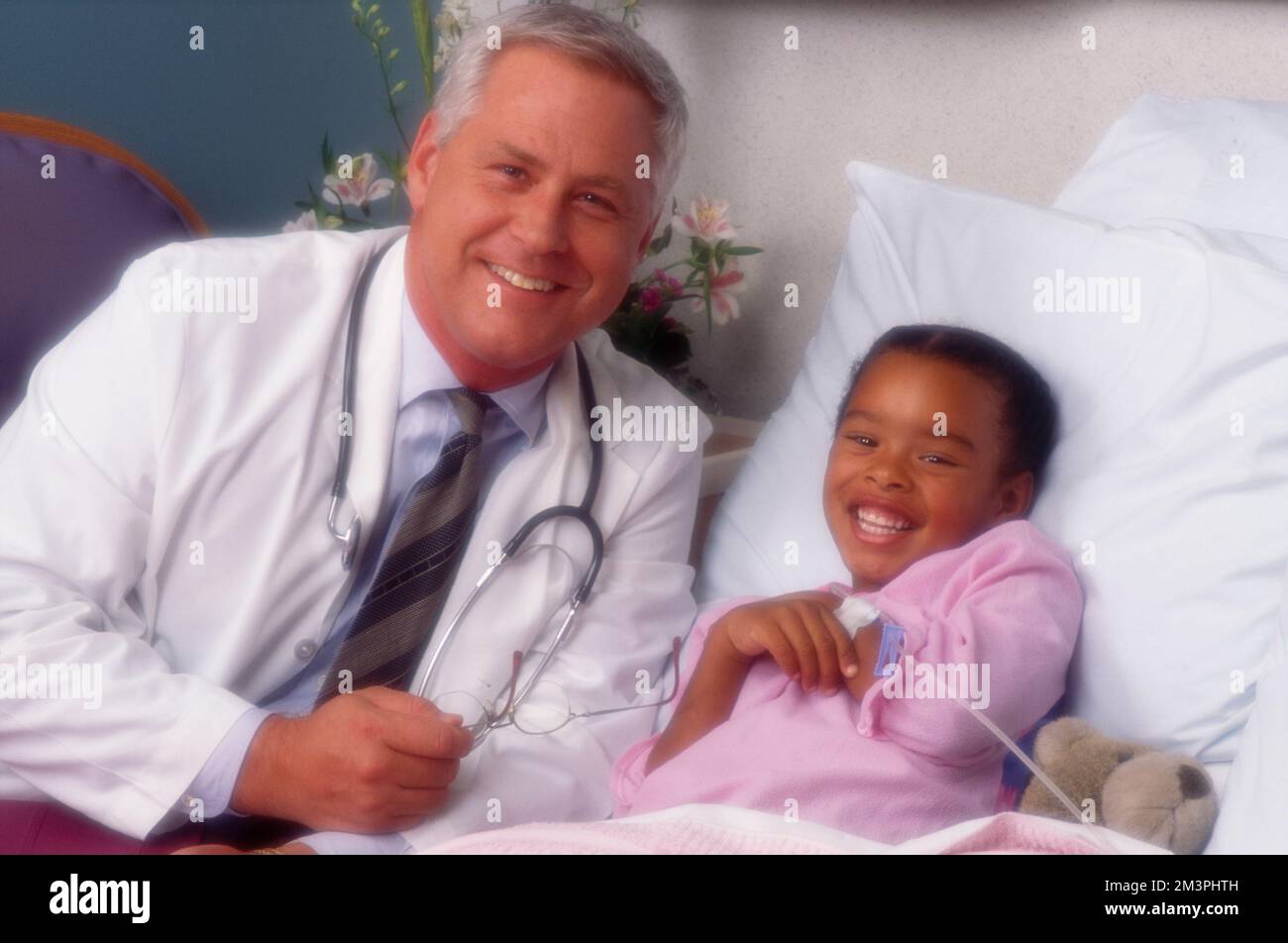 Older Male Doctor Sitting On The Edge Of The Bed Of A African American Girl Patient Both