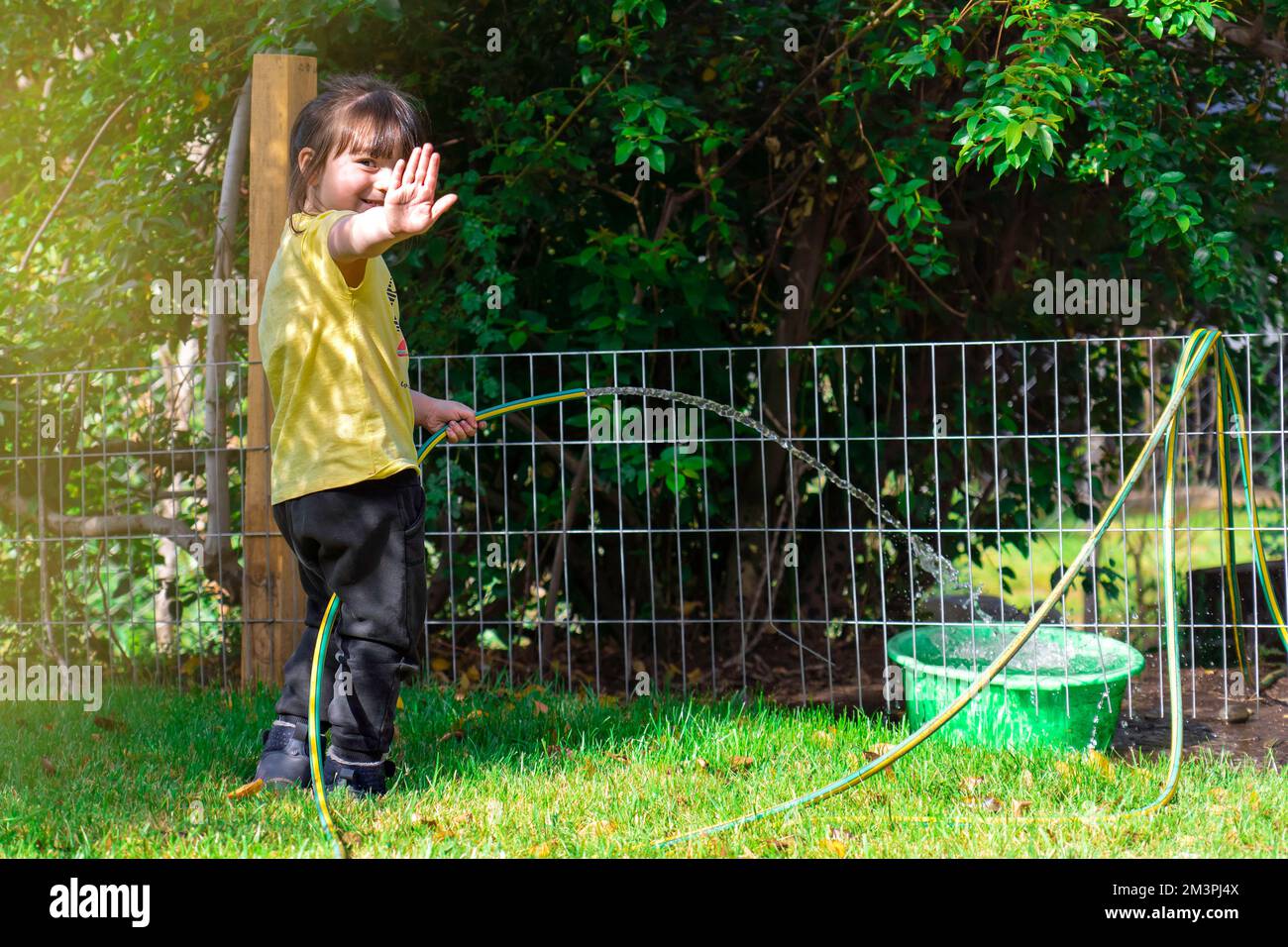 Happy little girl playing with a garden hose waving at the camera ...