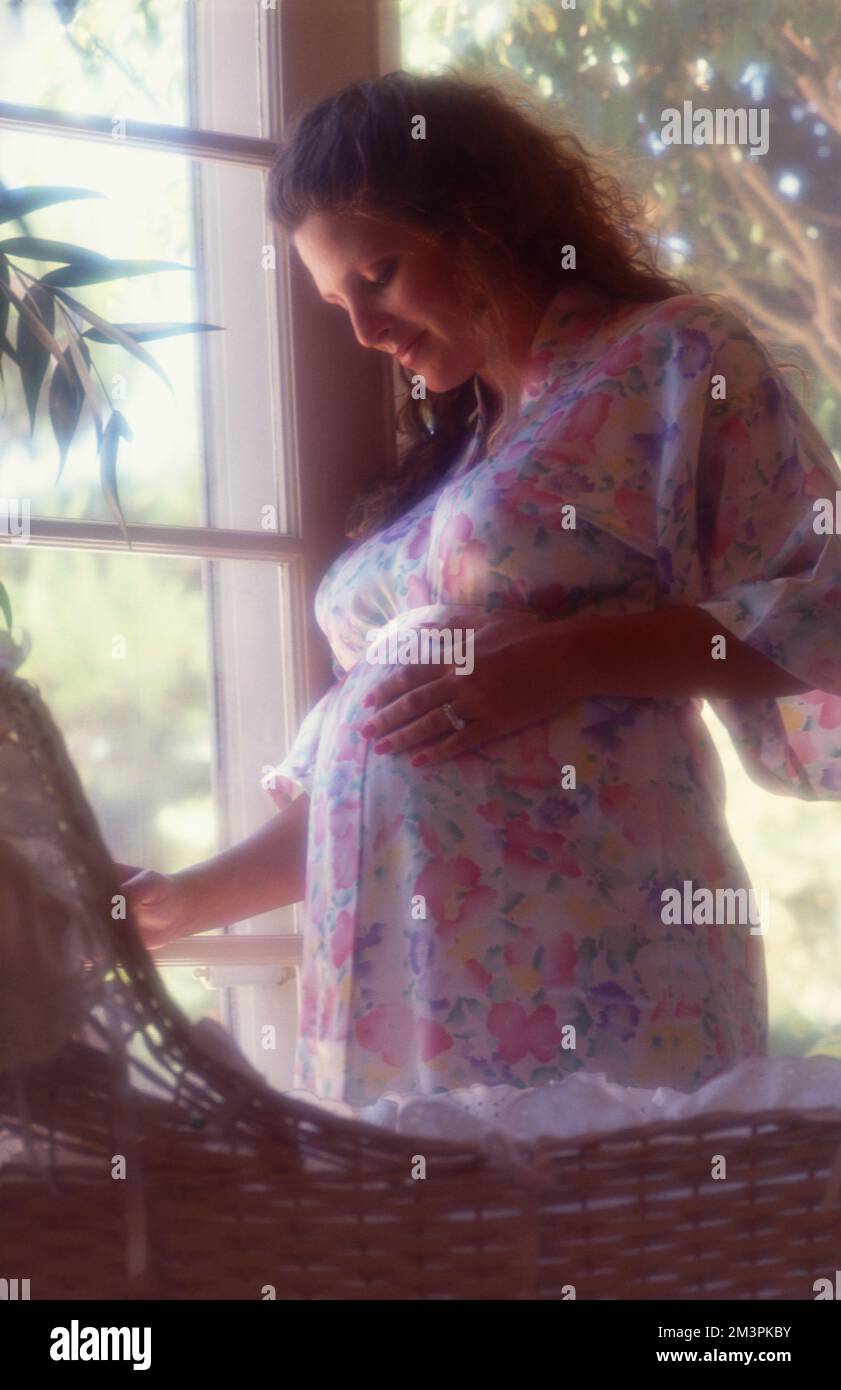 Pregnant woman in robe standing by a window hand on belly glancing down ...