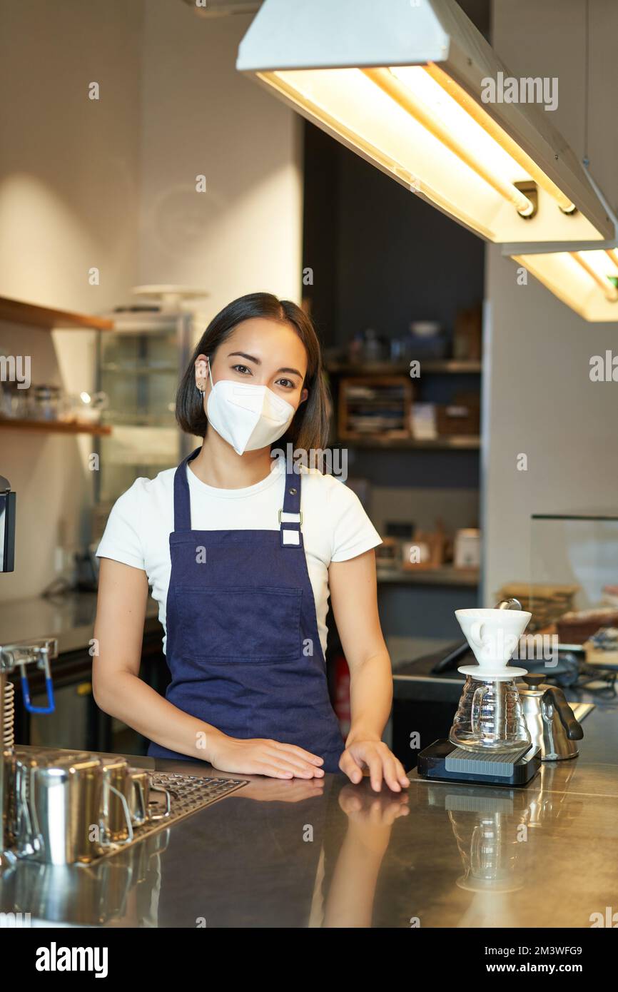 Vertical shot of friendly barista, female staff member in medical mask ...