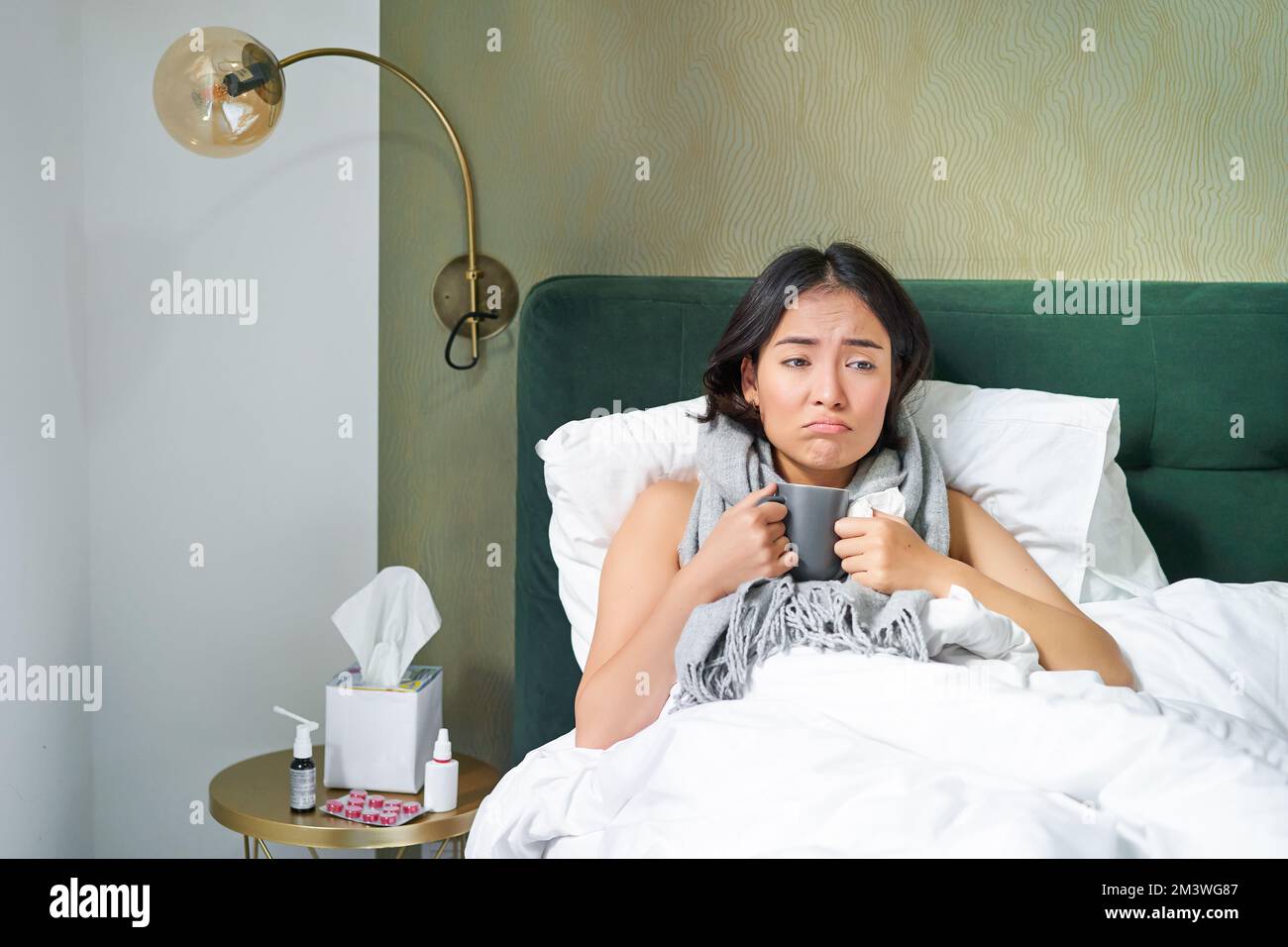 Sick sad asian girl lying in her bed with flu, catch a cold, taking pills and drinking hot tea ...