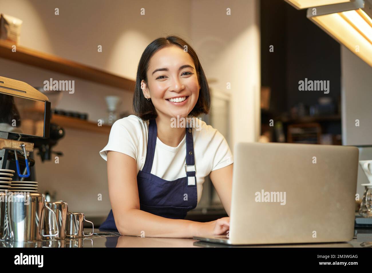 Enthusiastic asian girl in cafe uniform, barista worker with laptop ...
