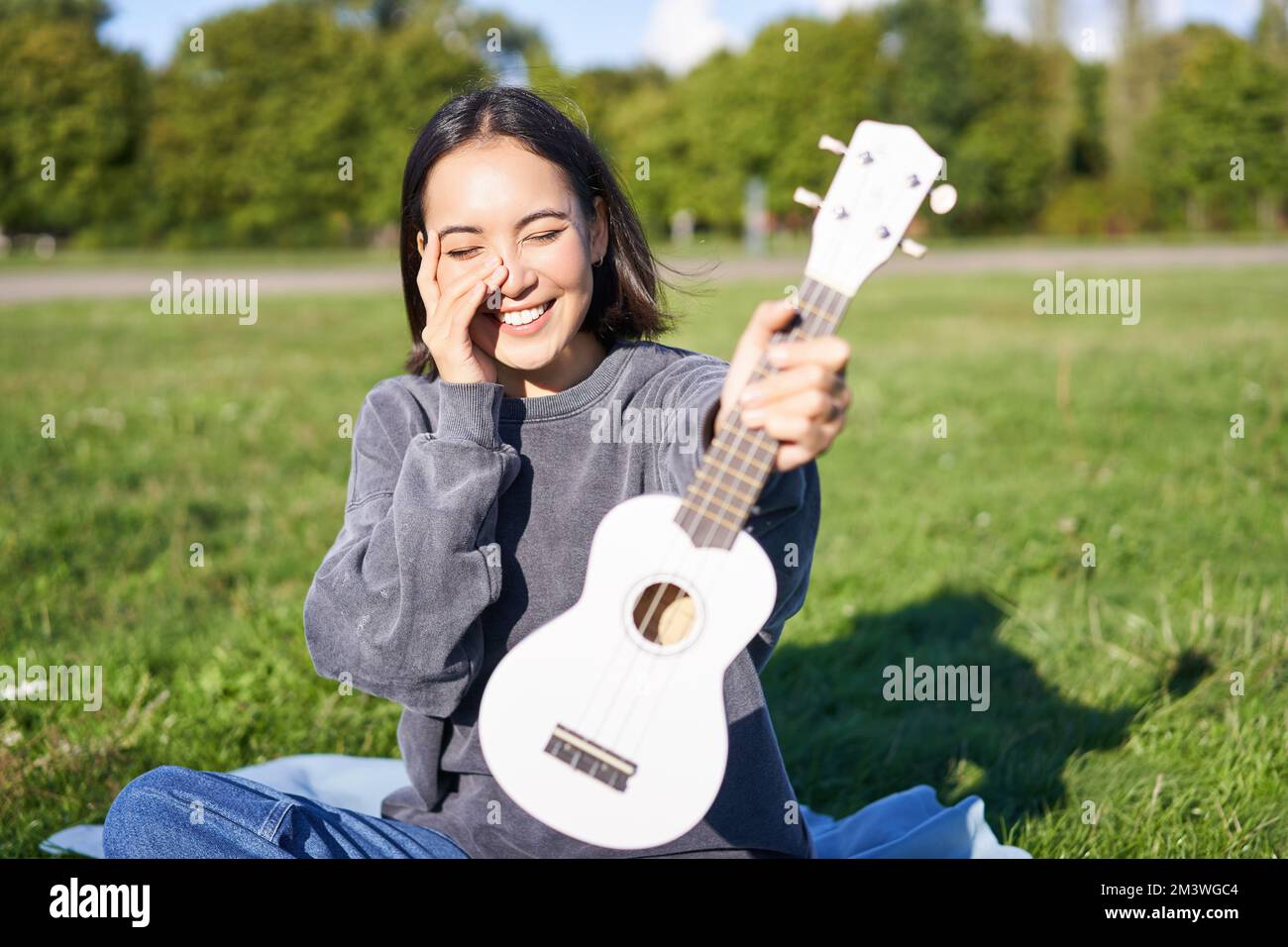 Beautiful asian girl with happy smile, shows her ukulele, sits outside ...