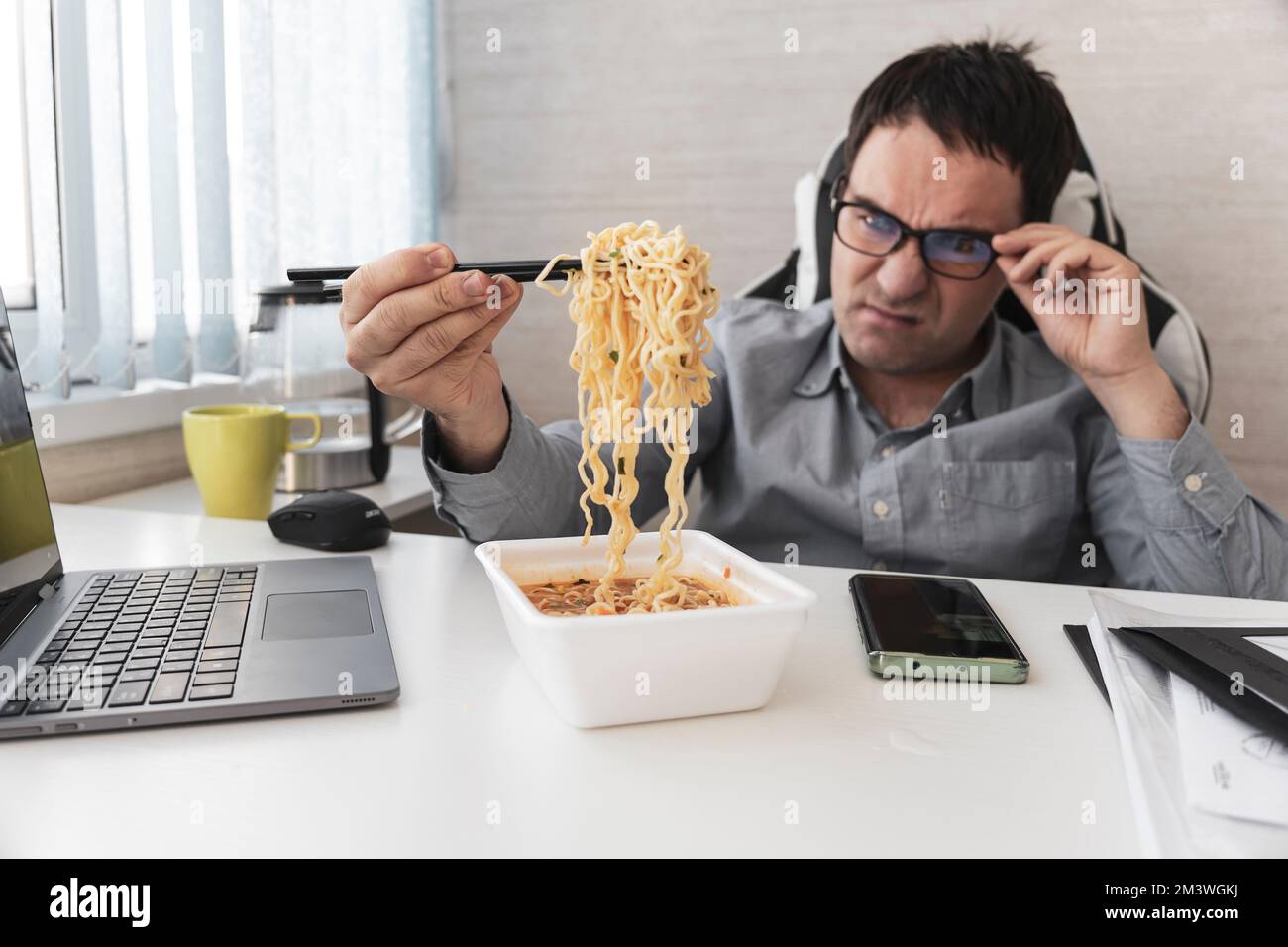 Young dissatisfied man eating instant noodles while working with laptop ...