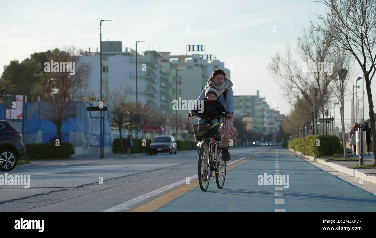 Mother rides bicycle with child in front in bike lane by sea shore ...