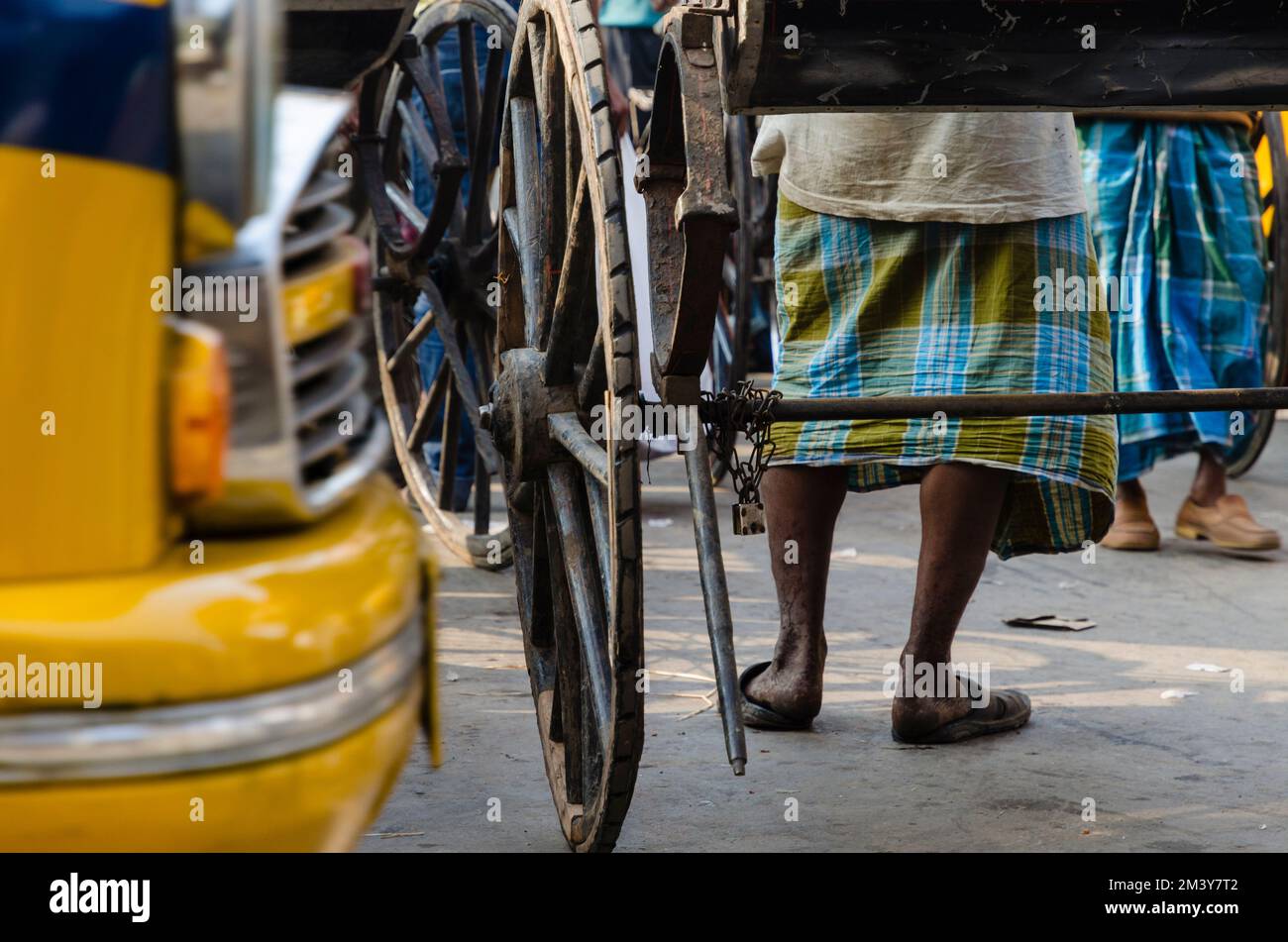 Wheel of a pull rikshaw and the legs of the kuli, yellow taxi next to ...