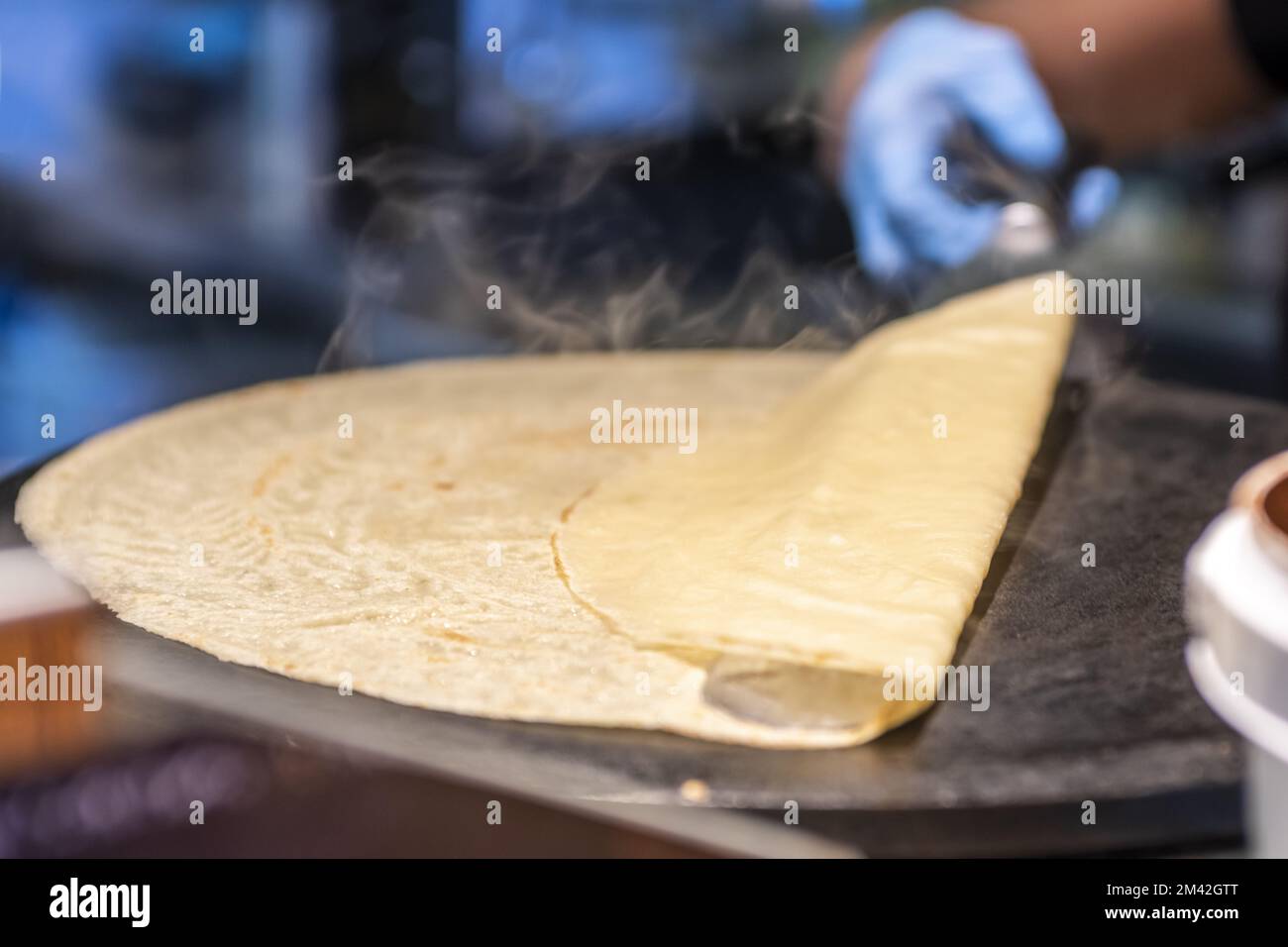A chef cooking a large French crepe blowing smoke on a metal griddle ...