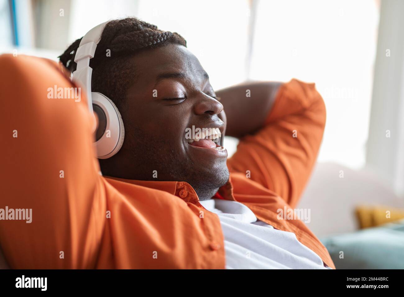 Positive black guy reclining on sofa, singing songs, using headphones ...