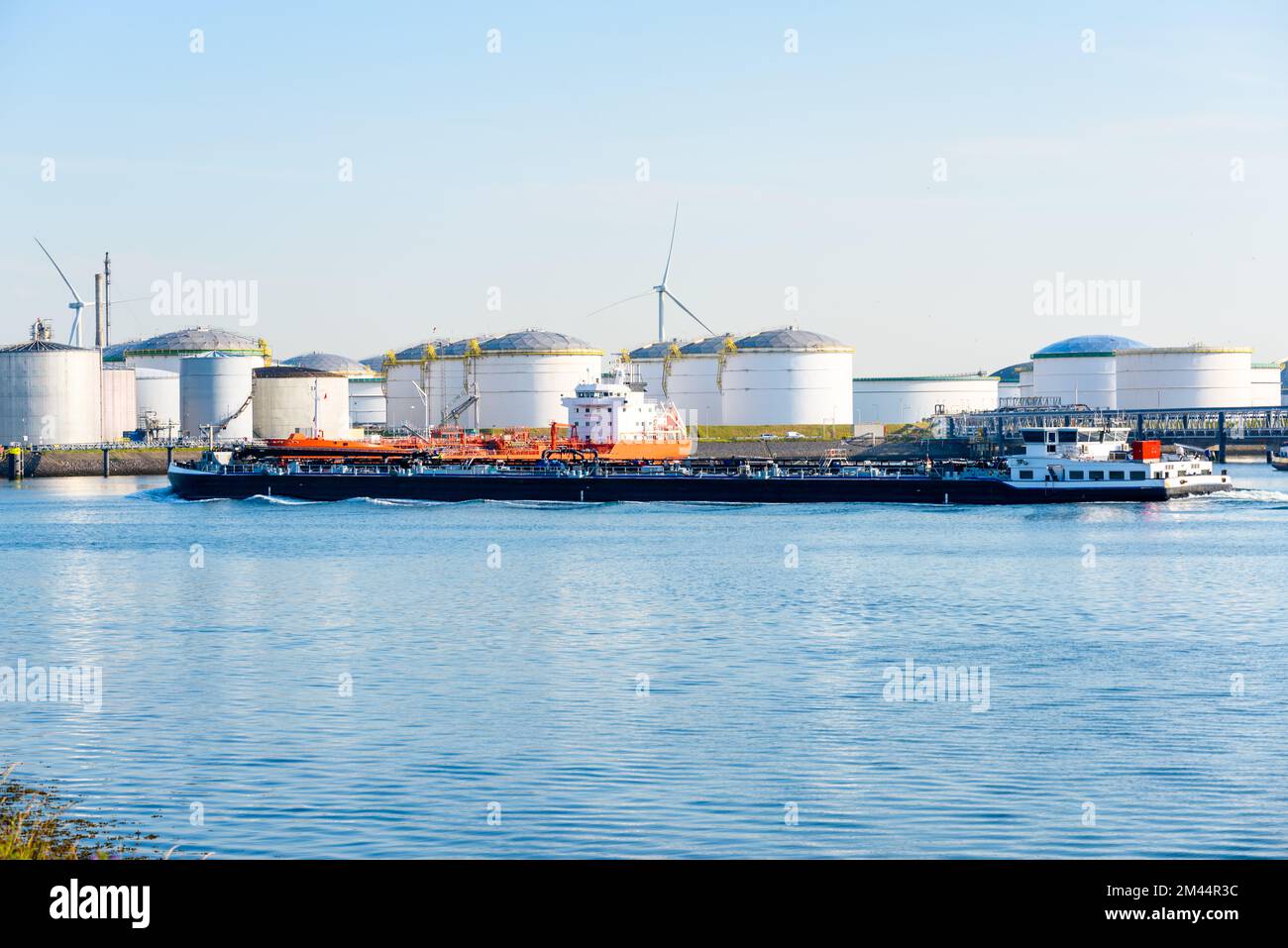 Tanker barge sailing past a oil termnal with large steel tanks on a ...