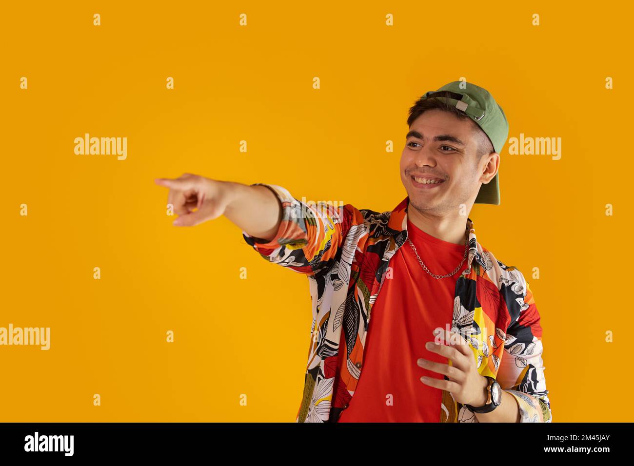 Young man in colourful shirt and cap pointing away against yellow ...