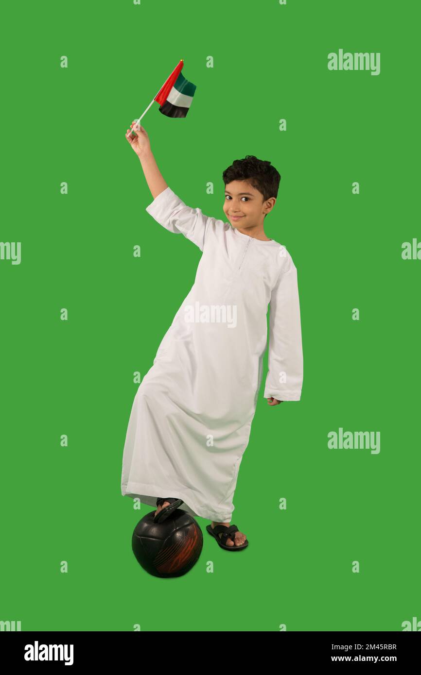 Arab boy holding a UAE national flag while one foot is on a football ...