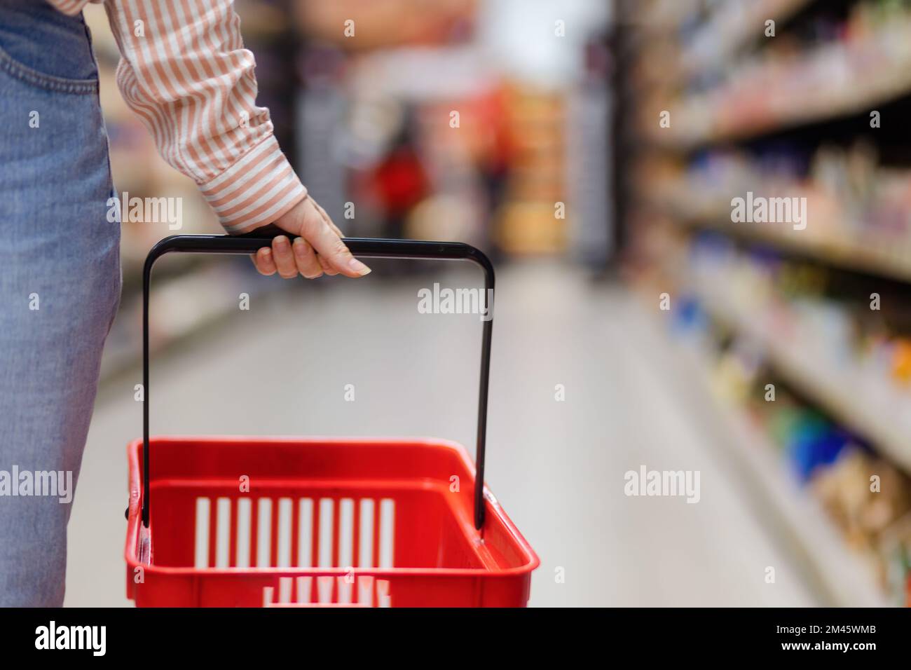 Female hand holds empty red grocery cart, close-up. In background is ...