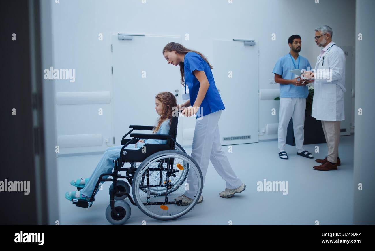 Young nurse pushing little girl on wheelchair at hospital corridor ...