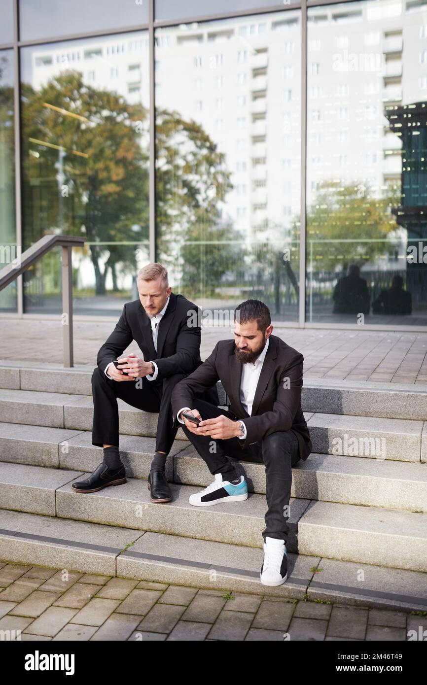 Businessmen sitting on stairs and using phones outside office building ...
