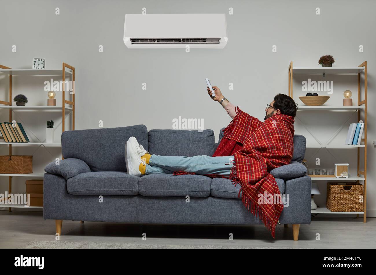 Young man wrapped in blanket holding air conditioner remote control ...