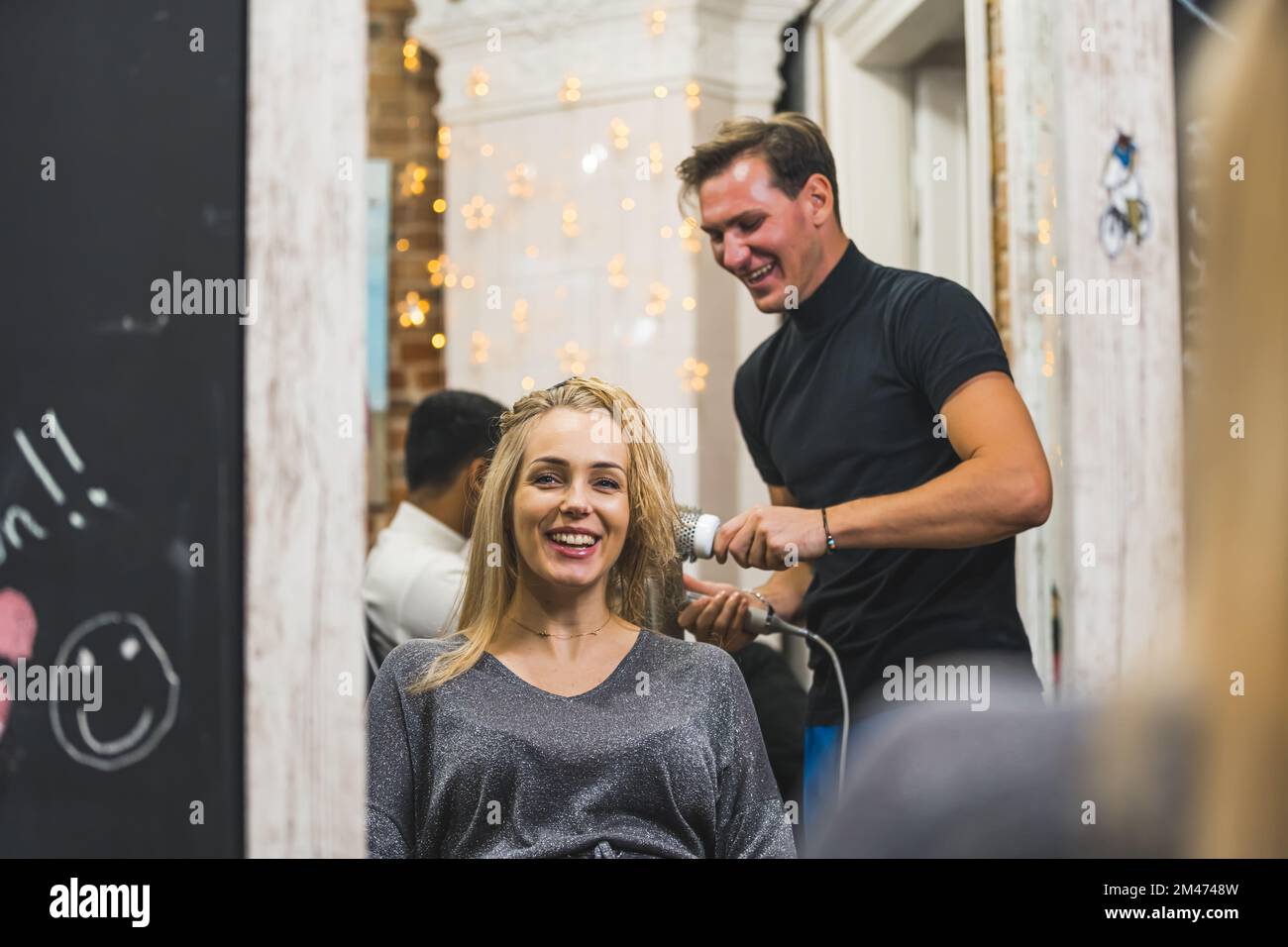 Mirror reflection of laughing adult woman on hairdresser chair having her hair styled by male ...