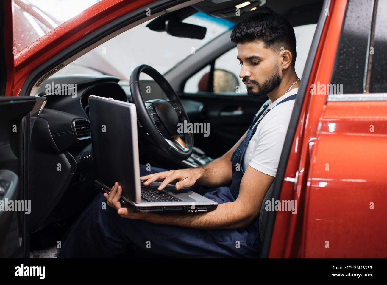 Bearded male mechanic sitting inside car using laptop, recording ...