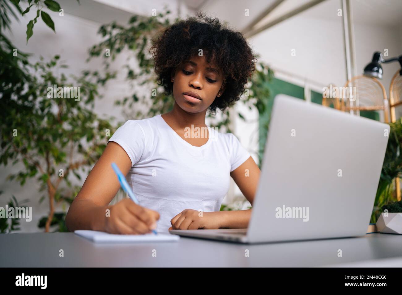 Low-angle view of focused African-American young woman writing information on notebook with pen ...