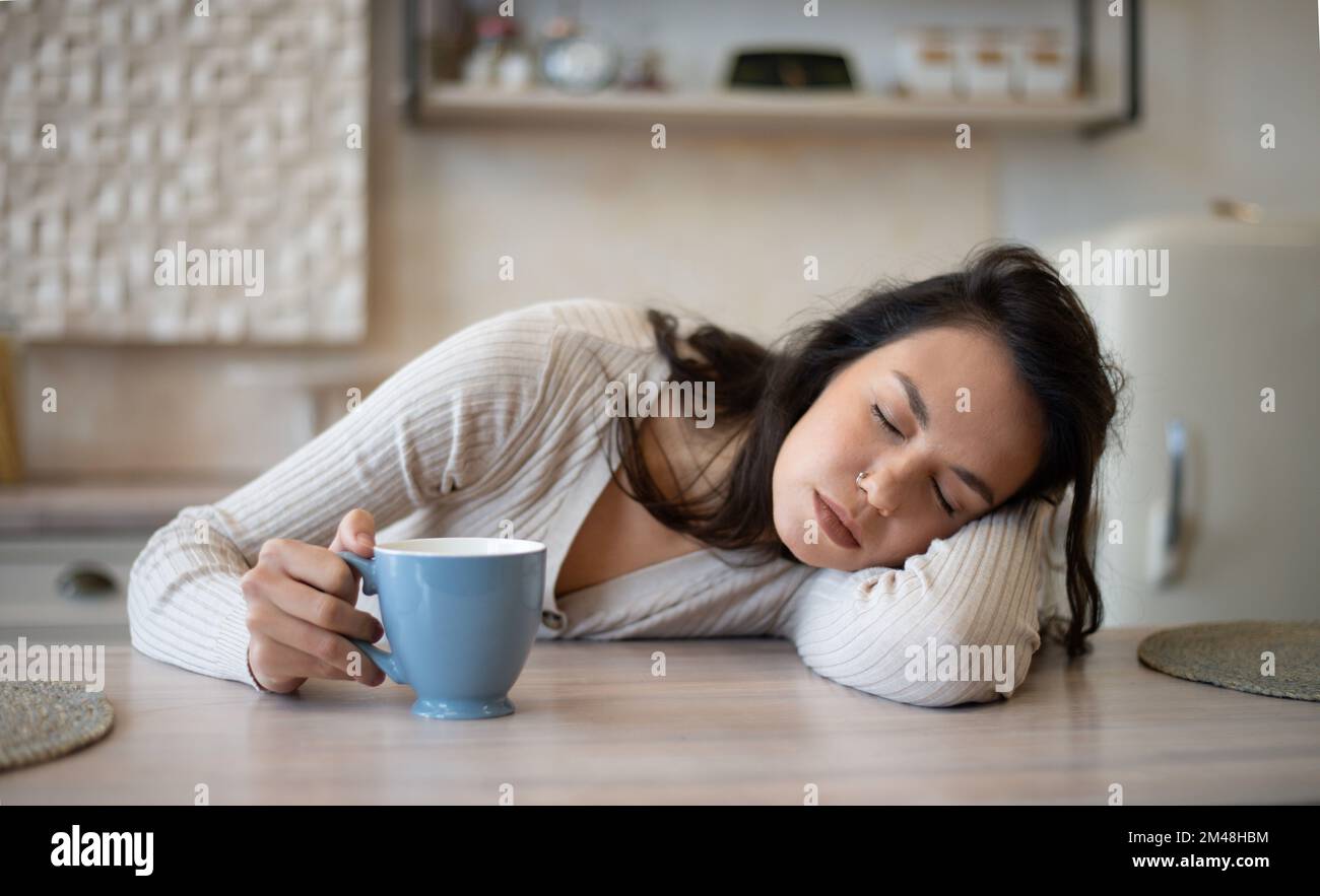 Tired woman with coffee cup sleeping on table in modern light kitchen ...