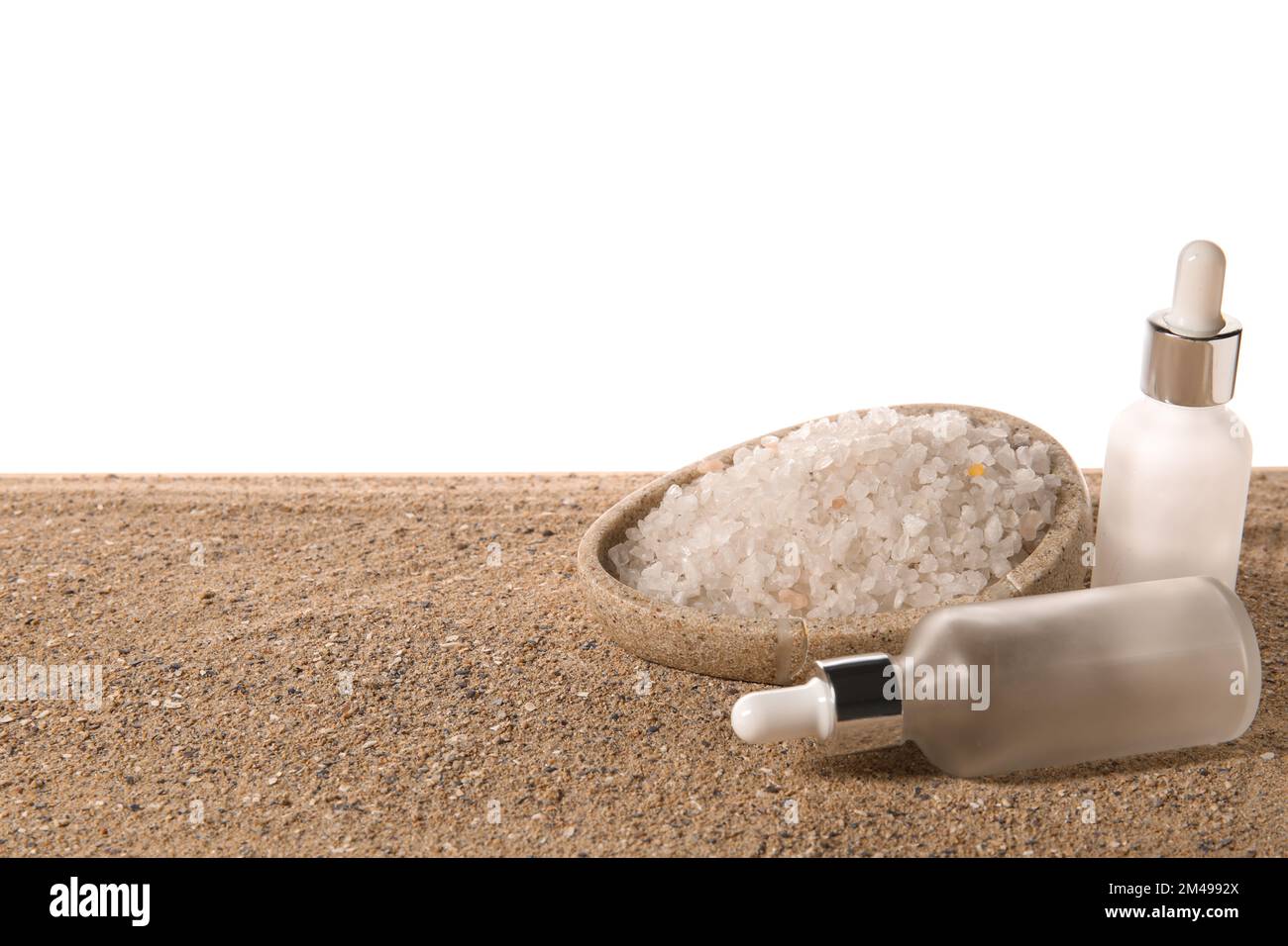 Cosmetic dropper bottles with bowl of sea salt on sand against white ...
