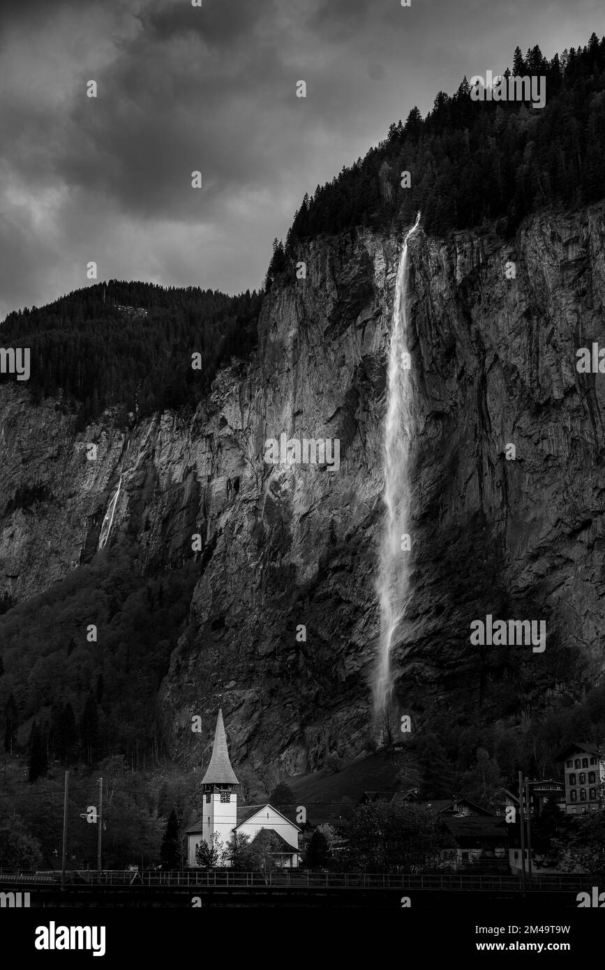 A grayscale of a church and waterfall flowing from a cliff with trees ...