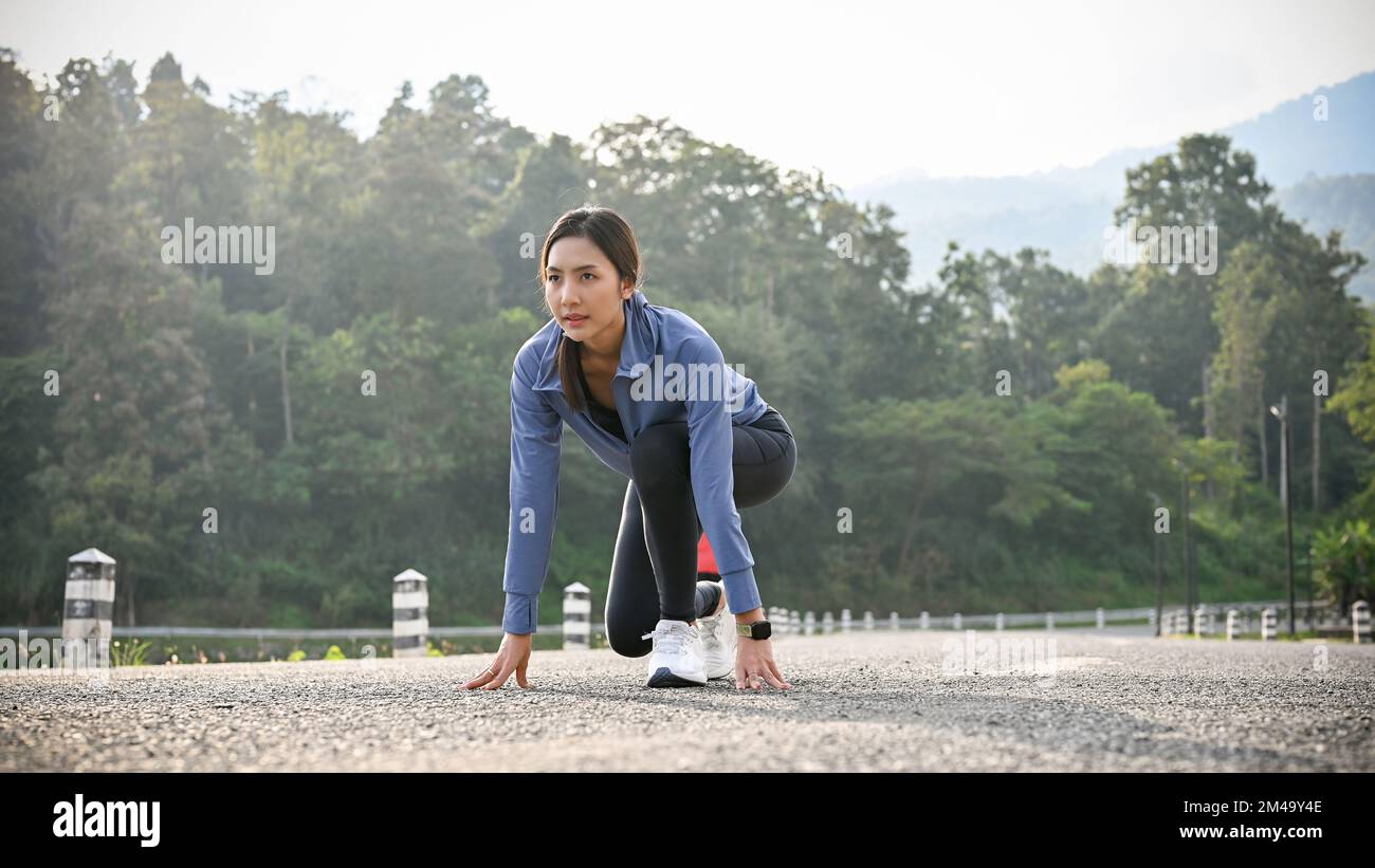Determined and focused Asian female runner in sportswear in running start pose, preparing to run ...