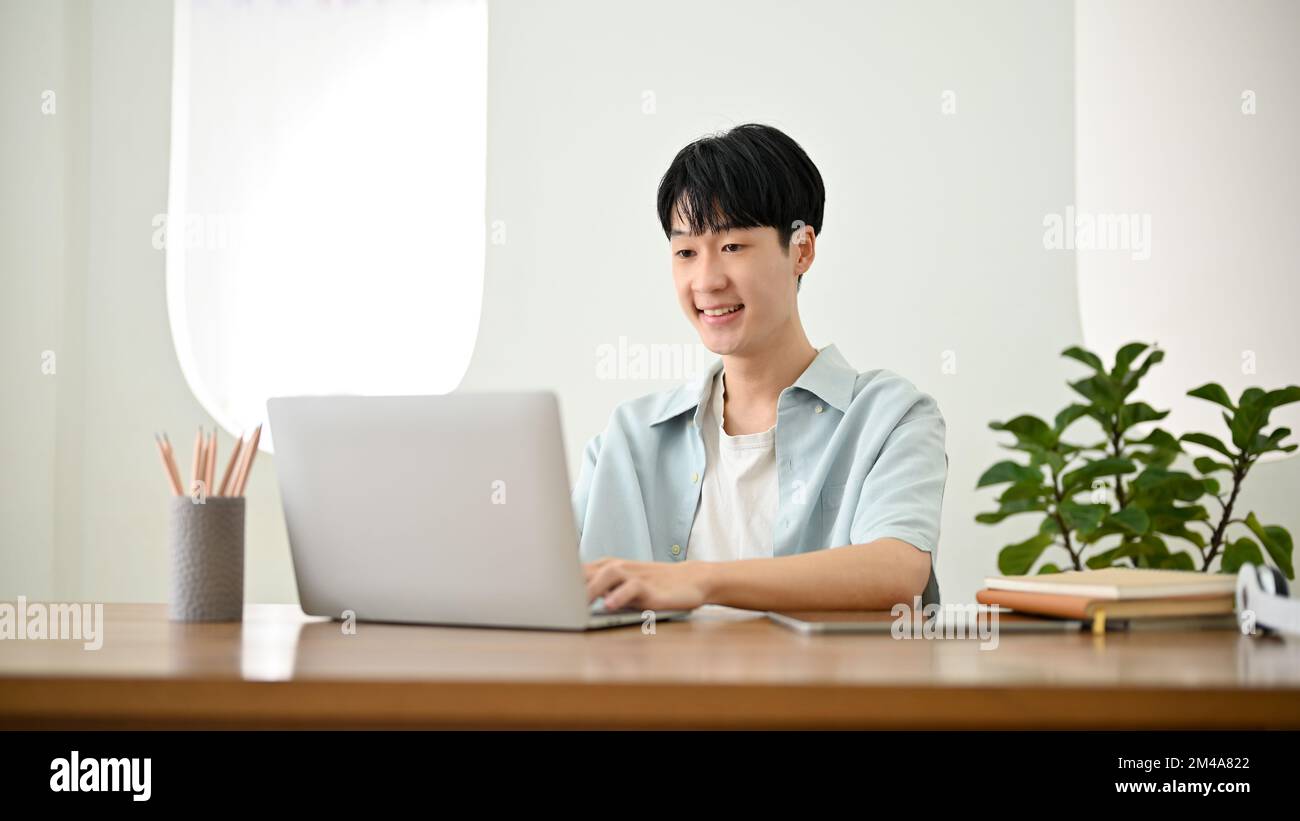 Happy and handsome young Asian man working on his tasks on laptop in ...