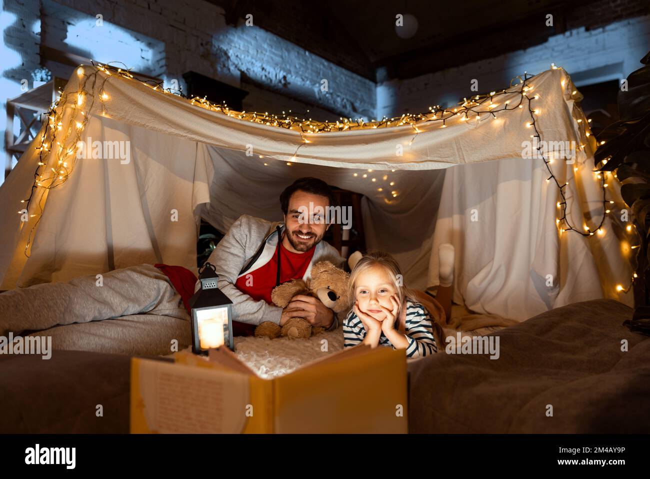 Cheerful father and daughter lying inside self-made hut, tent in room ...