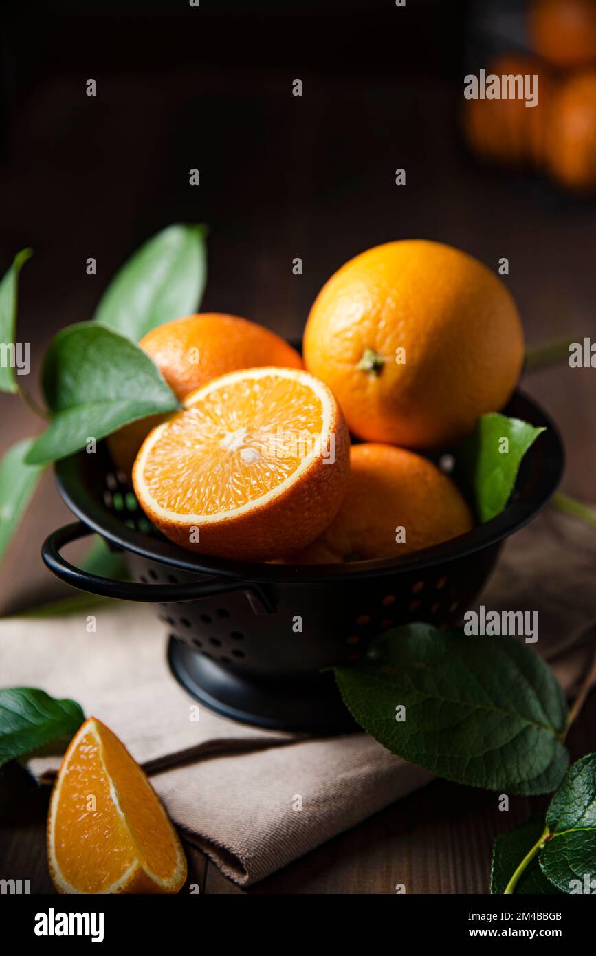 A few juicy oranges in a black bowl with green leaves on a dark wooden ...