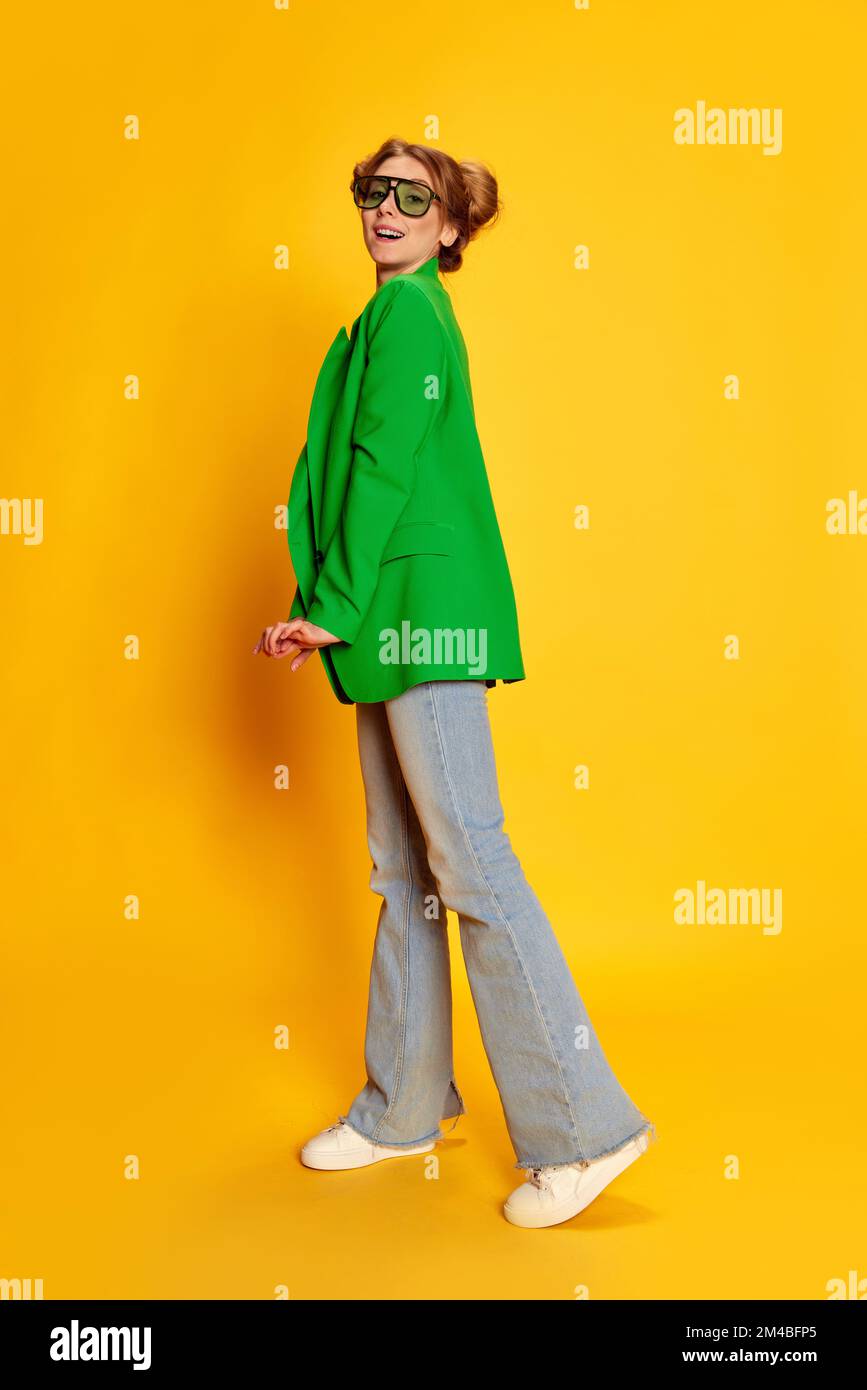 Portrait of young girl in bright green jacket posing over yellow studio ...