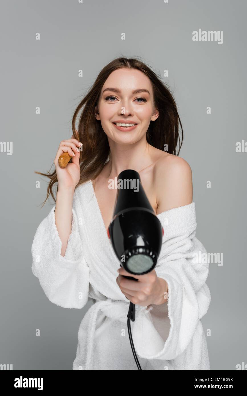 smiling young woman with shiny hair using hair dryer and brushing hair ...