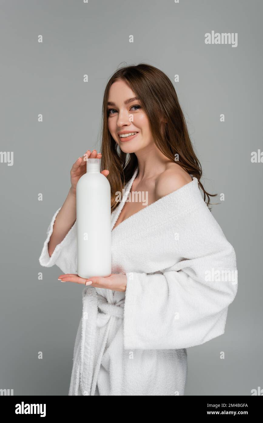 joyful young woman with shiny hair holding bottle with shampoo isolated ...