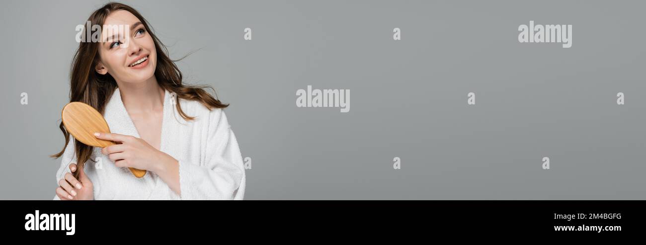 pleased young woman holding wooden hair brush while brushing shiny hair ...