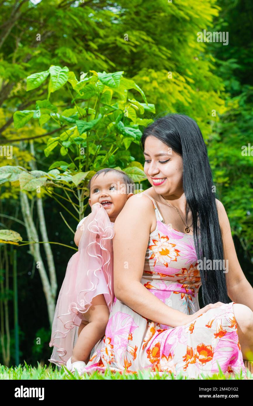 very happy Latina mother, sitting on the grass, next to her little ...