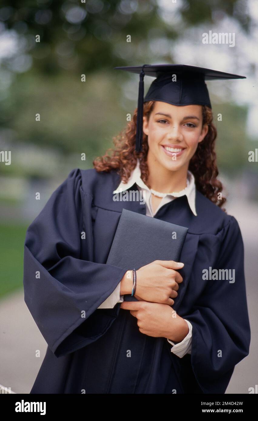 African American girl posing in cap and gown and holding her graduation certificate, she smiling ...