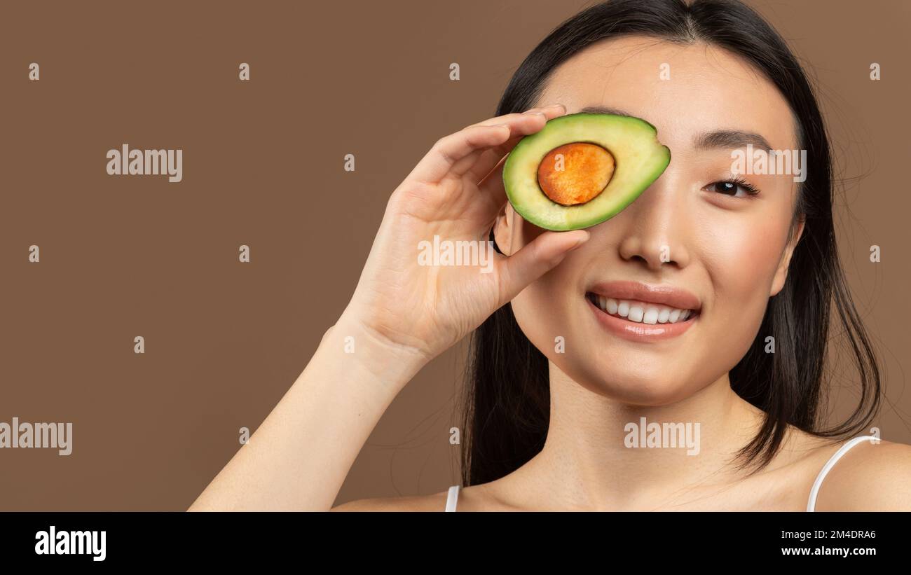 Portrait of young asian woman holding avocado hald and covering eye ...