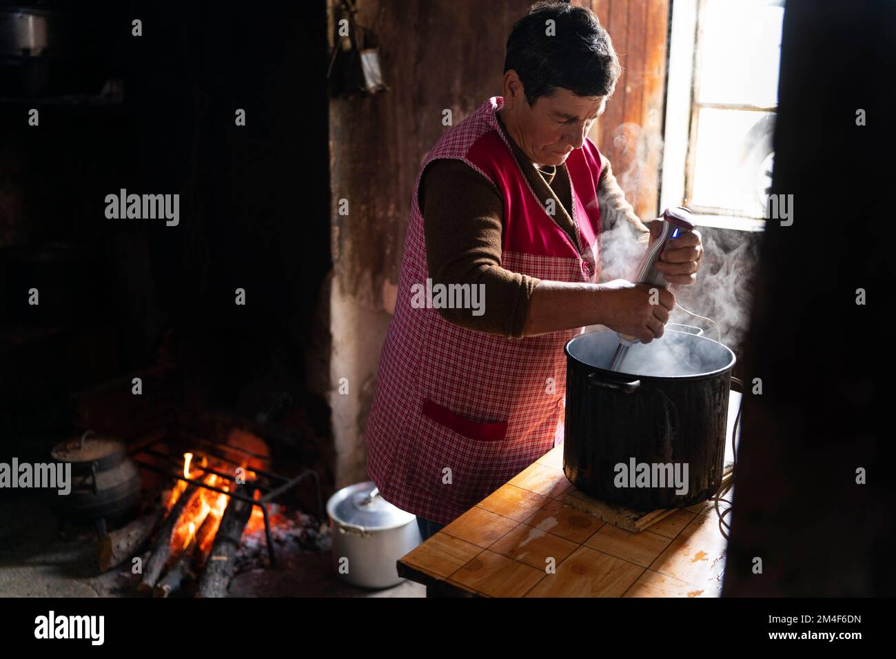 Old woman cooking on open fire in a small dimly lit kitchen in a ...