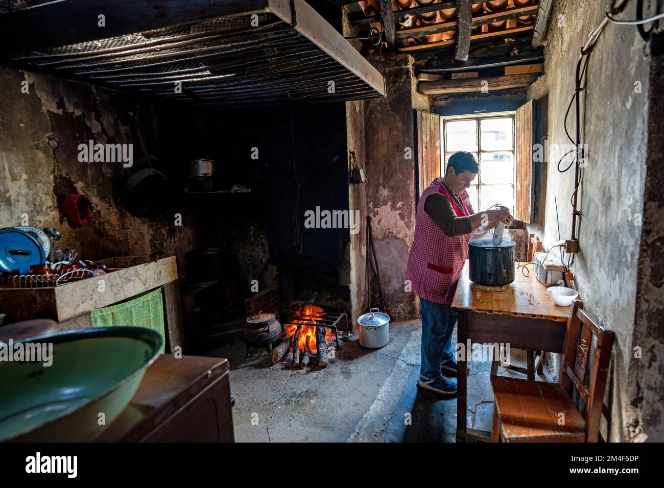 Old woman cooking on open fire in a small dimly lit kitchen in a ...