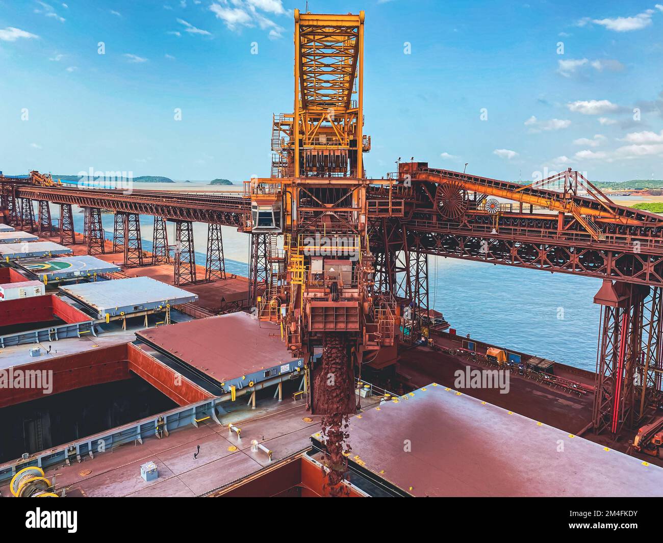 An Iron Ore Conveyor Loading Cargo In The Bulk Carrier With A Cloudy Blue Sky In The Background