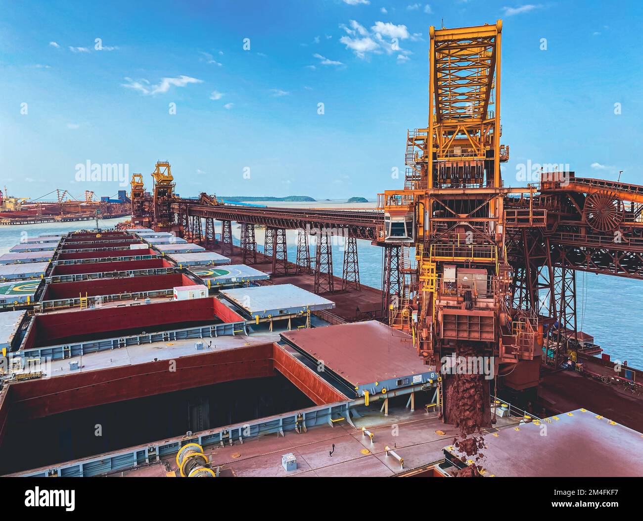 An Iron Ore Conveyor Loading Cargo In The Bulk Carrier With A Cloudy Blue Sky In The Background