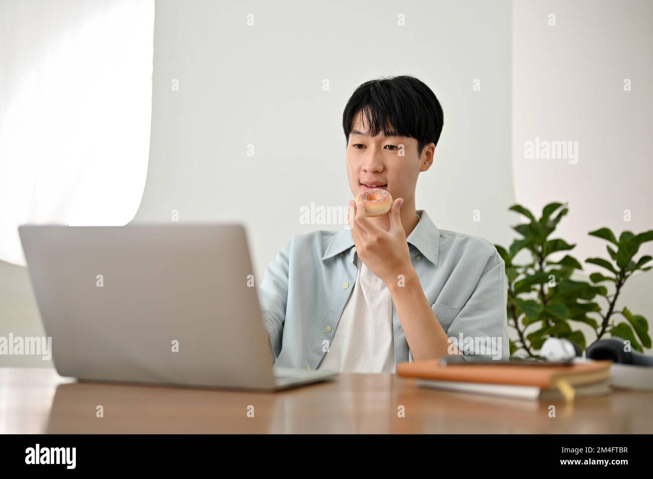 Handsome young Asian male college student eating donut while working on ...
