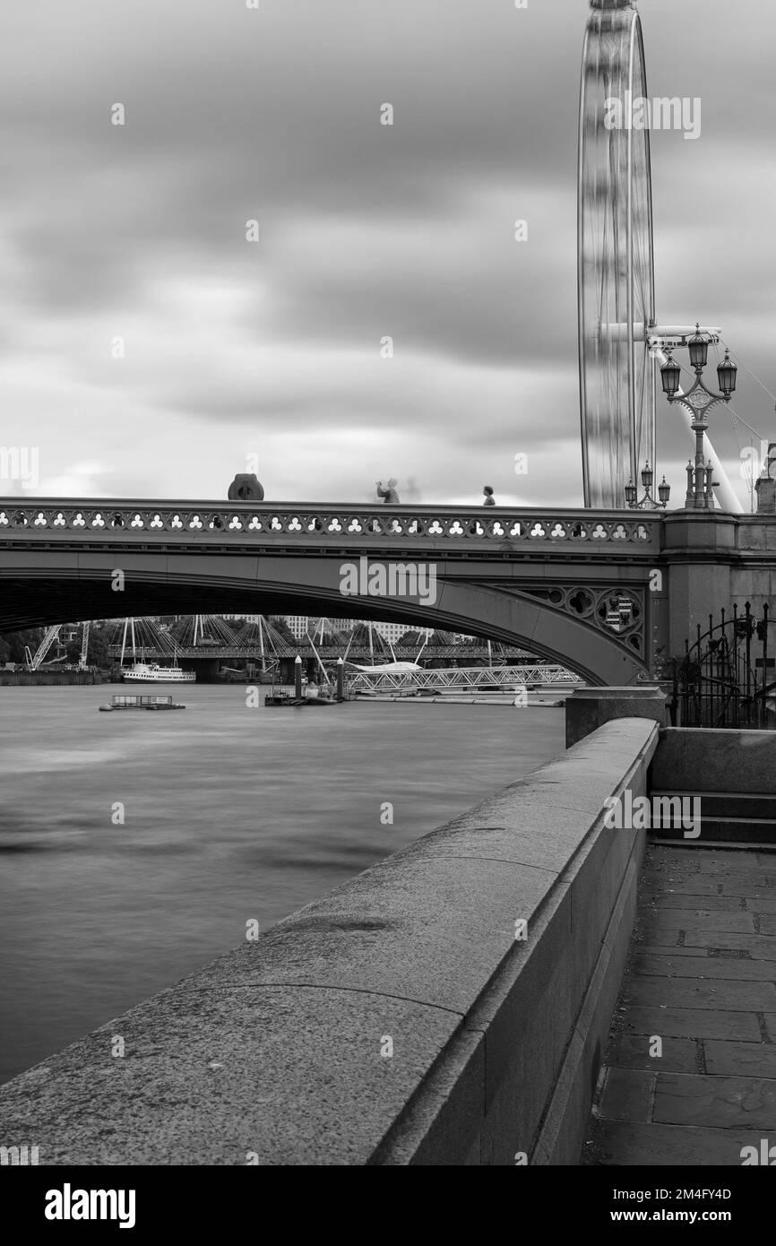 A vertical grayscale shot of a bridge with the background of the London ...