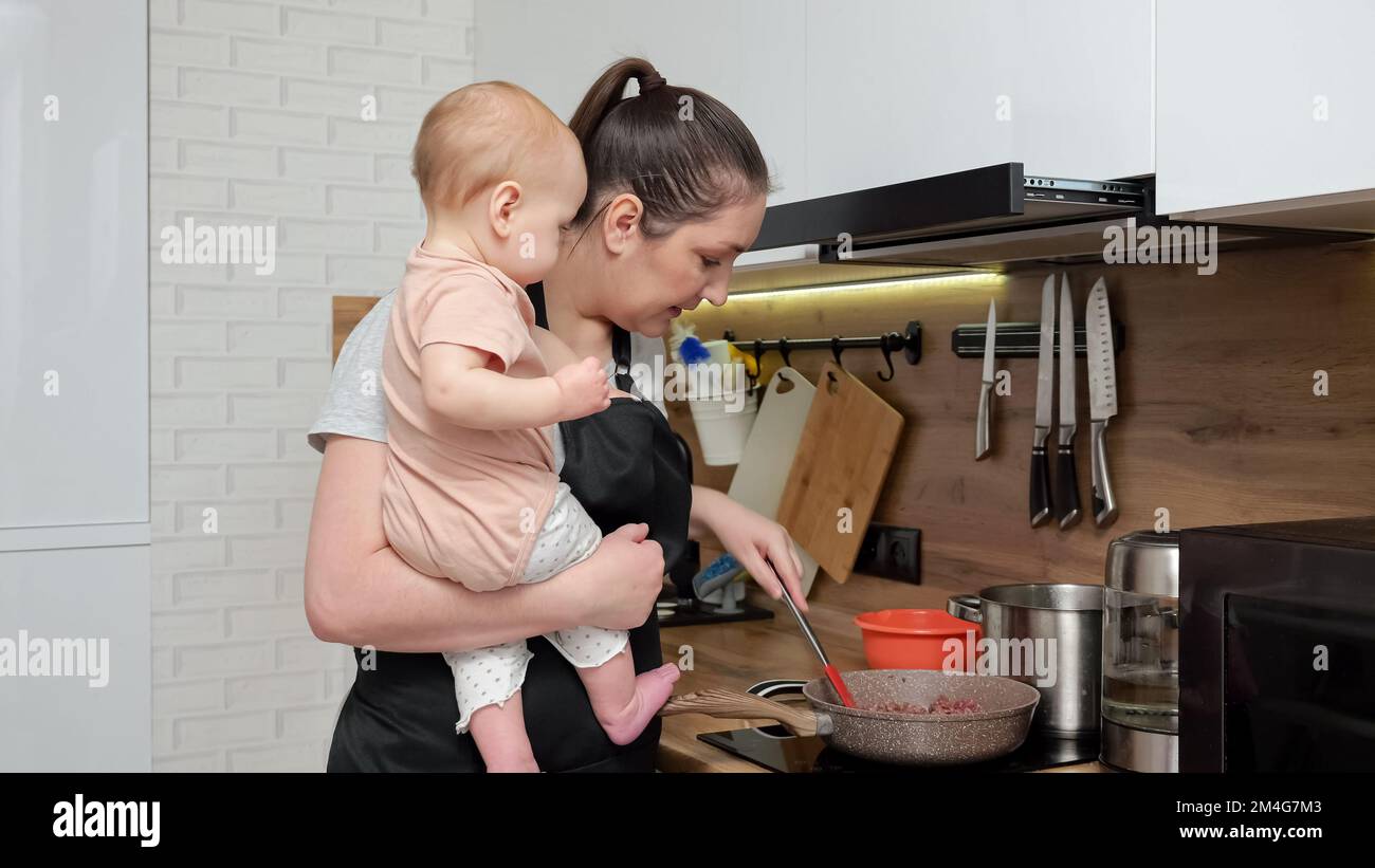 Blonde baby girl looks at busy mother cooking food in equipped kitchen ...
