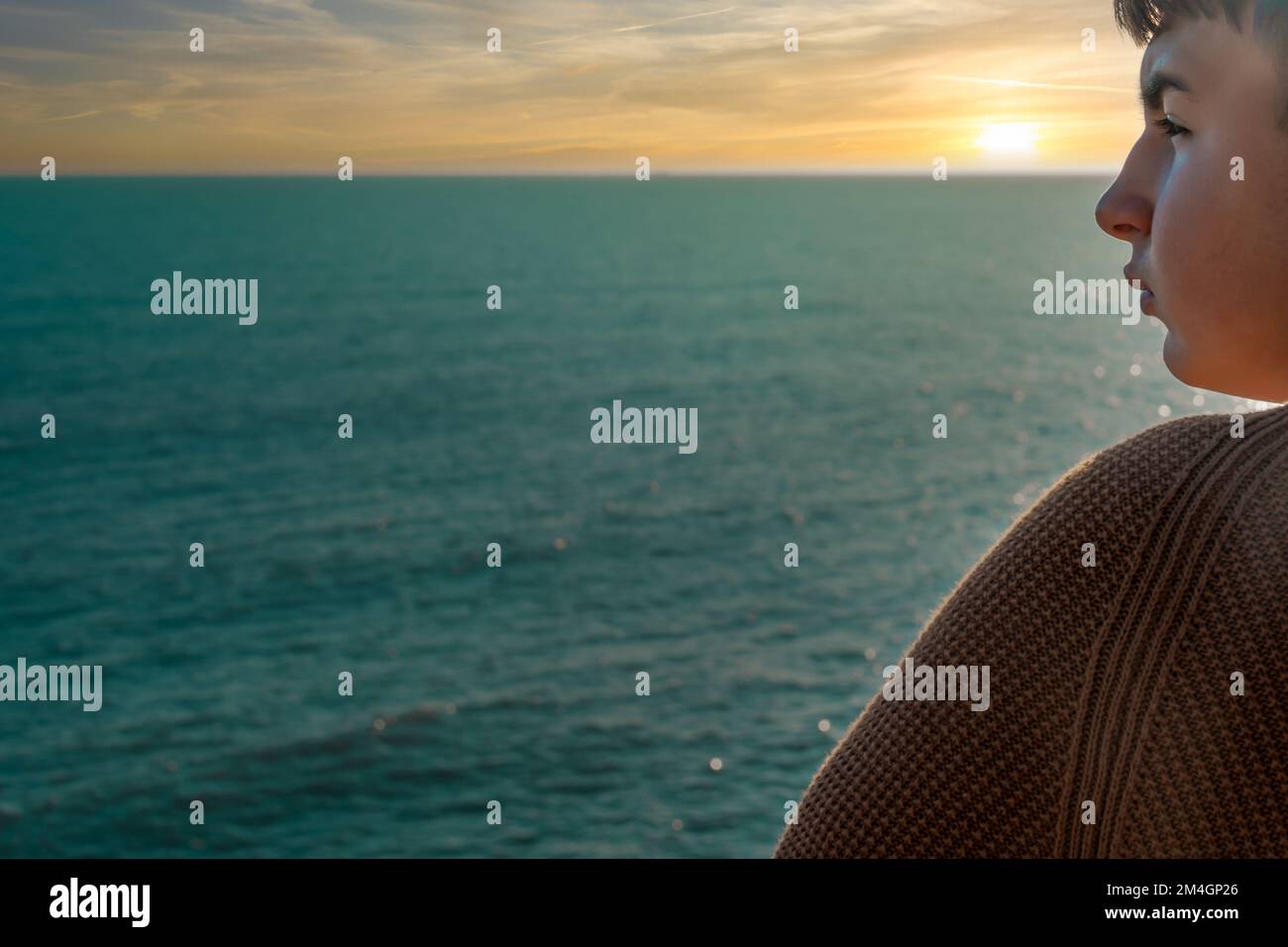 Teen boy in sweater looking away against waving sea and sundown sky in ...