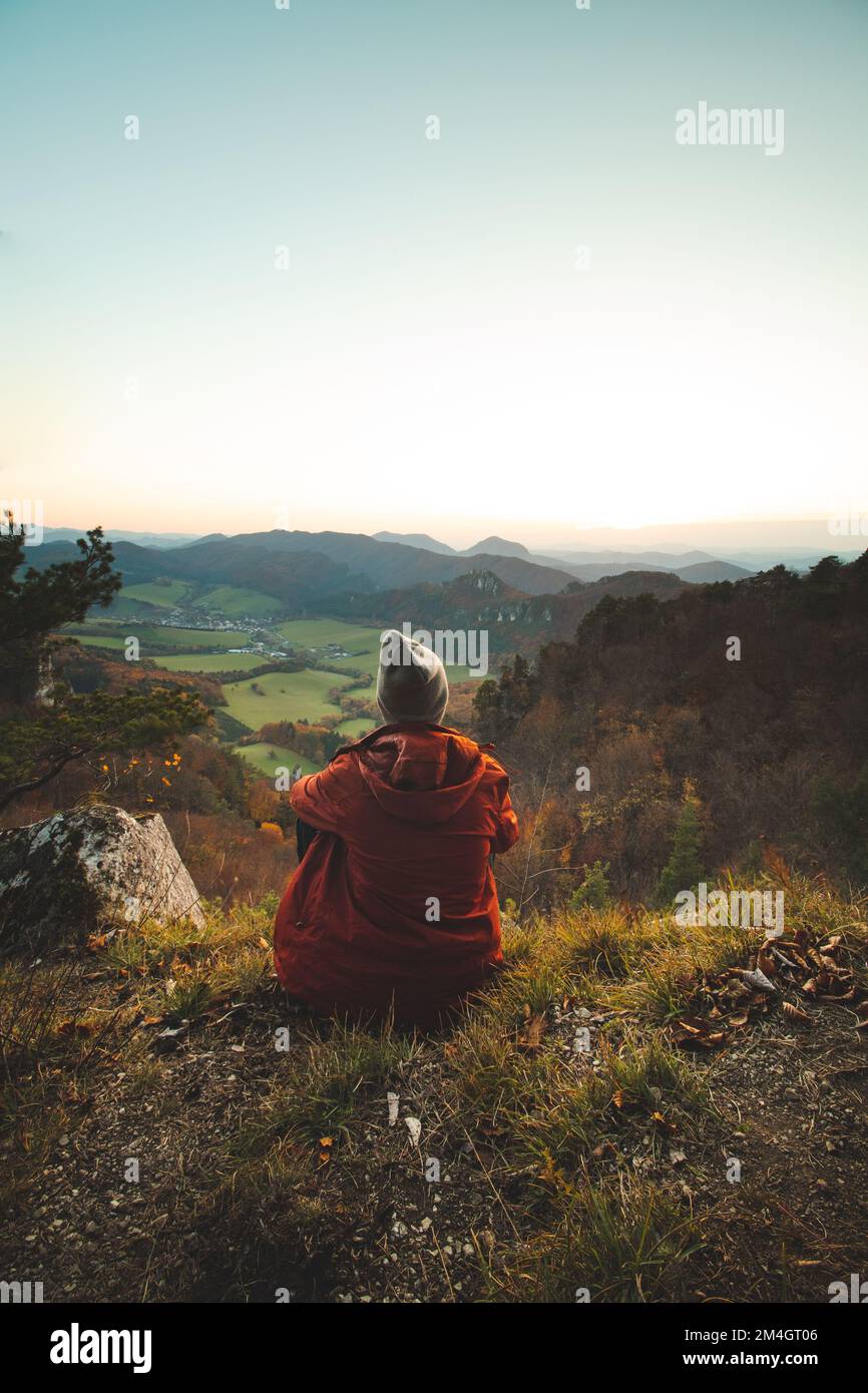 Enthusiastic traveller sitting on the edge of a rock in a red jacket ...