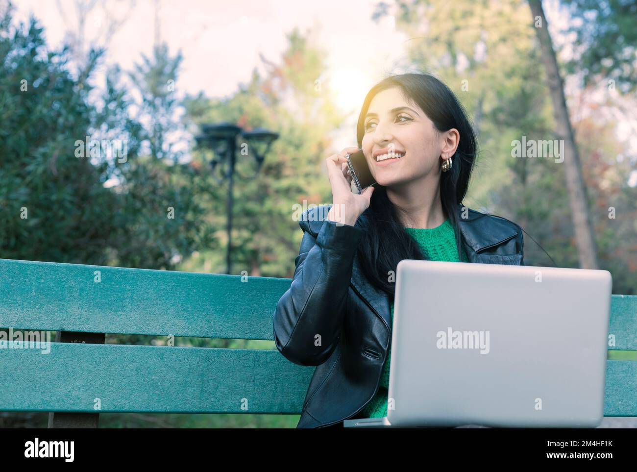 Young smiling fun student freelancer woman 20s in green jacket jeans sit on bench in spring park ...