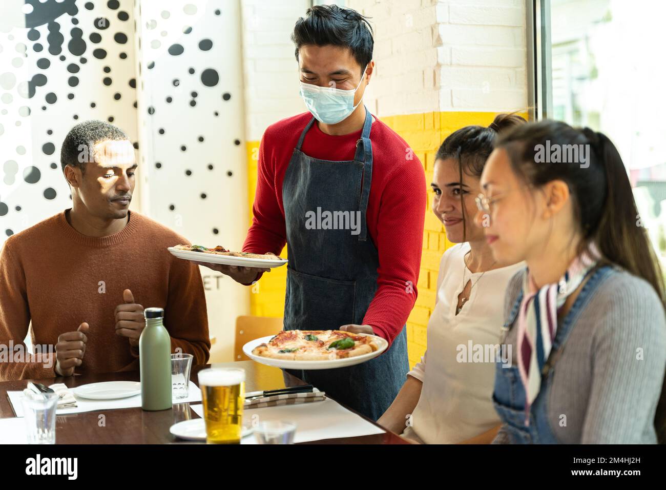 Smiling waiter with a protective face mask serving delicious pizza to a ...